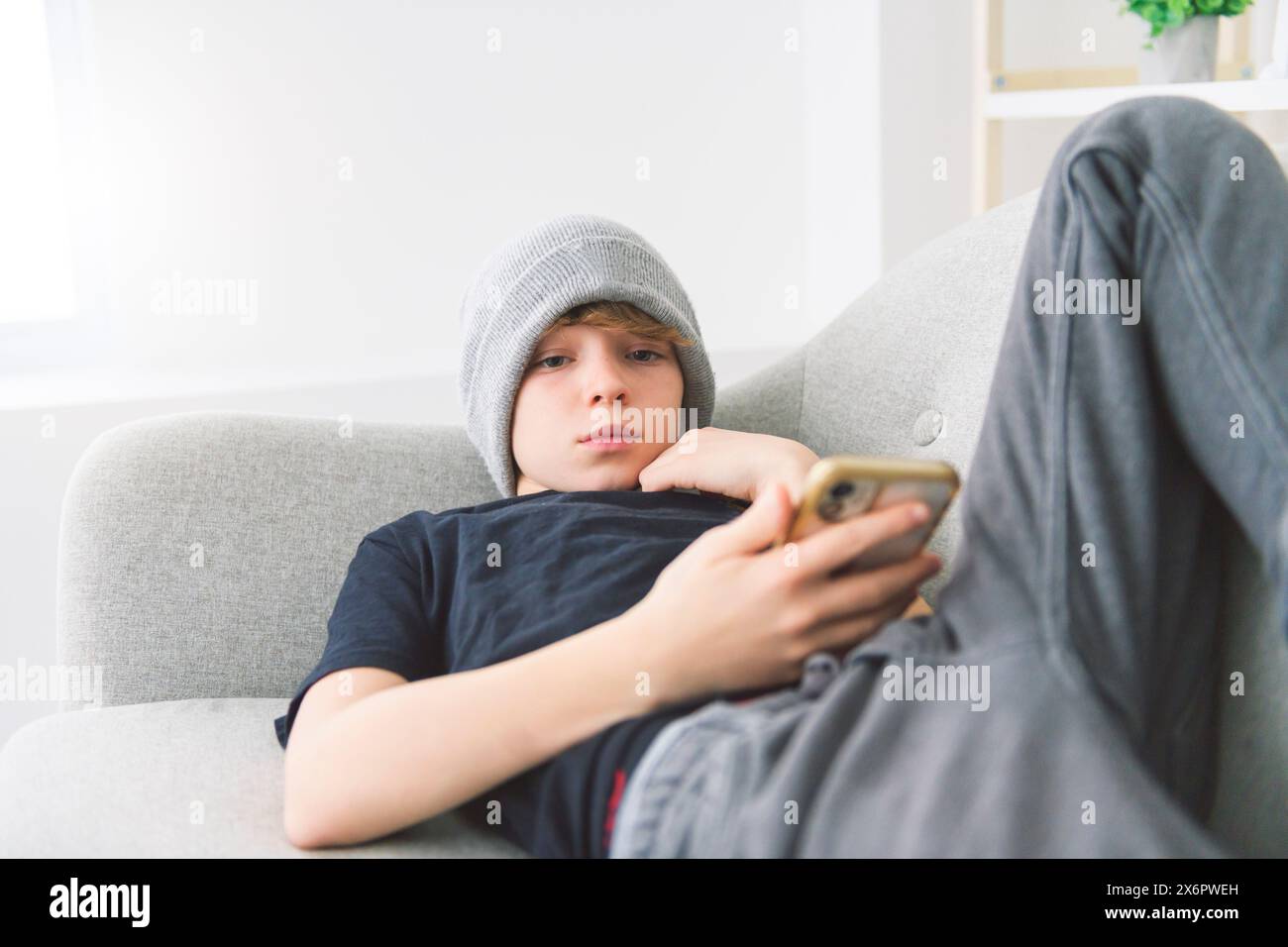 young boy lay on the couch at home watching cellphone wearing toque ...
