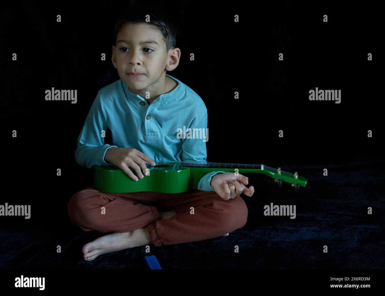 latino child sitting cross-legged on the floor holding a green ukulele ...