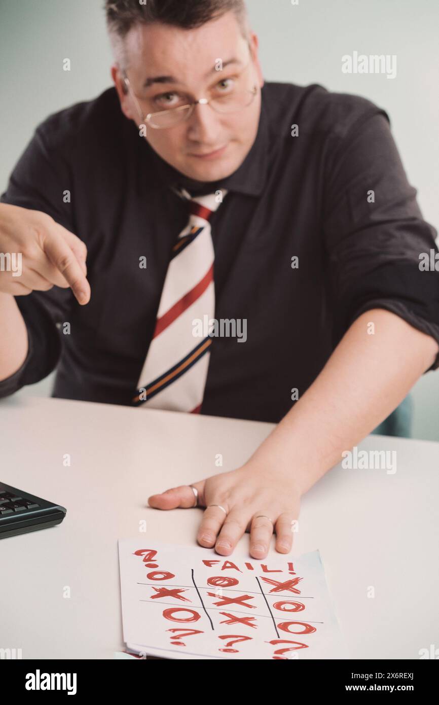 Seated man in glasses, dark shirt, and striped tie points at a tic-tac ...