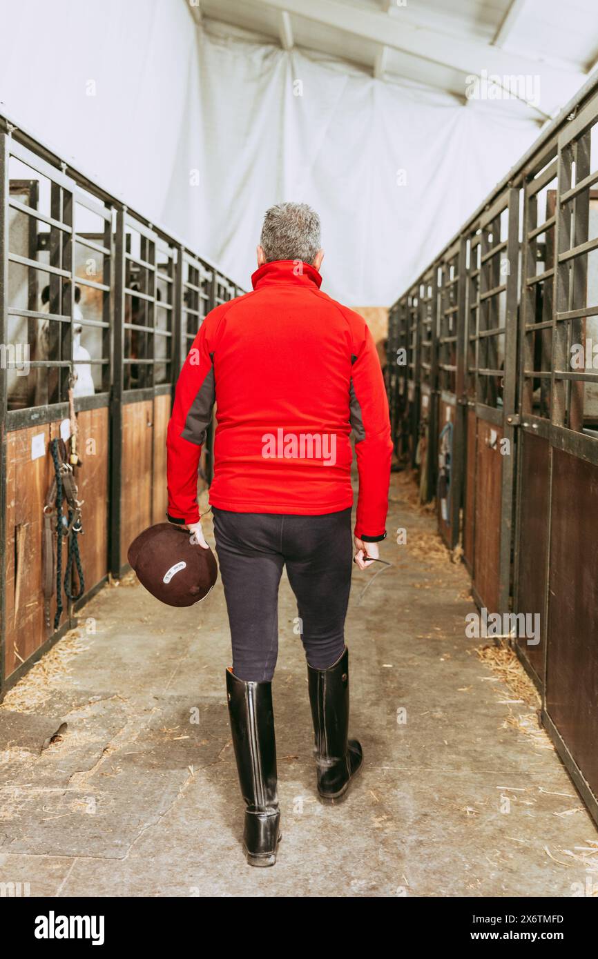 Back view of an equestrian walking down the stable aisle, holding a ...