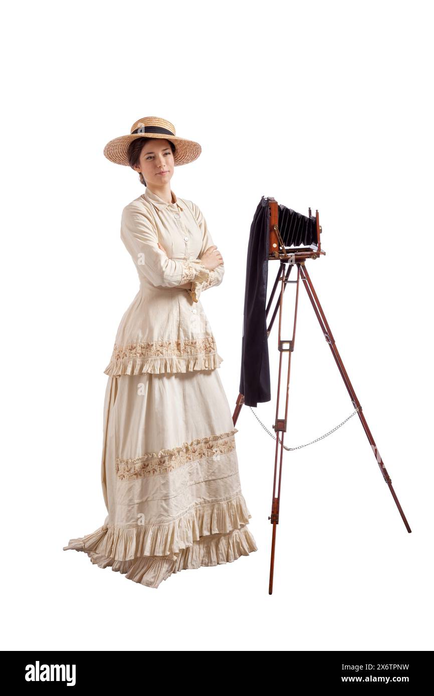 Young woman in Victorian dress with a straw hat standing by a camera on ...