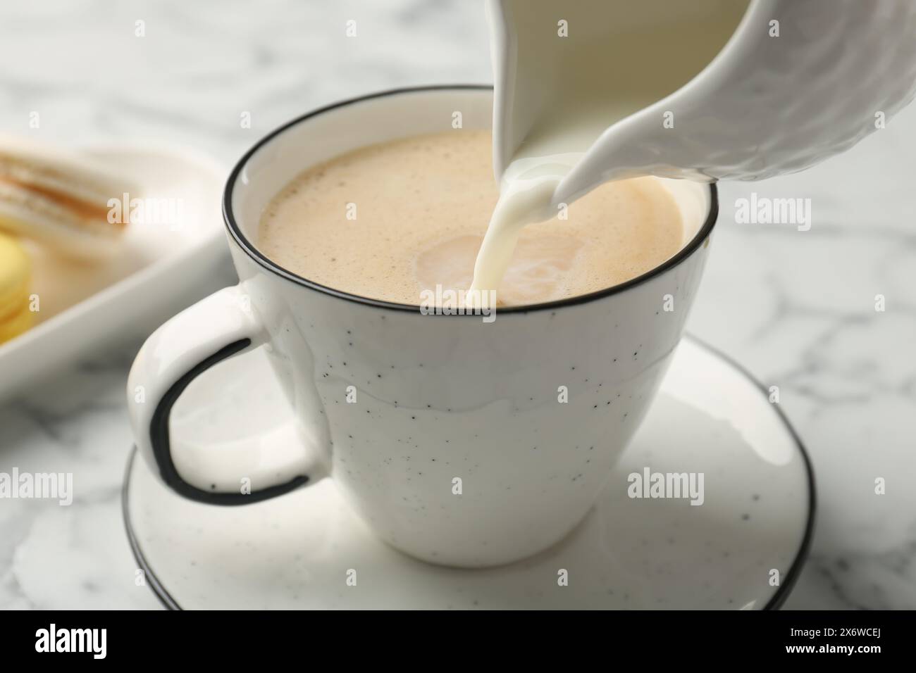 Pouring milk from pitcher into cup with coffee at white marble table ...