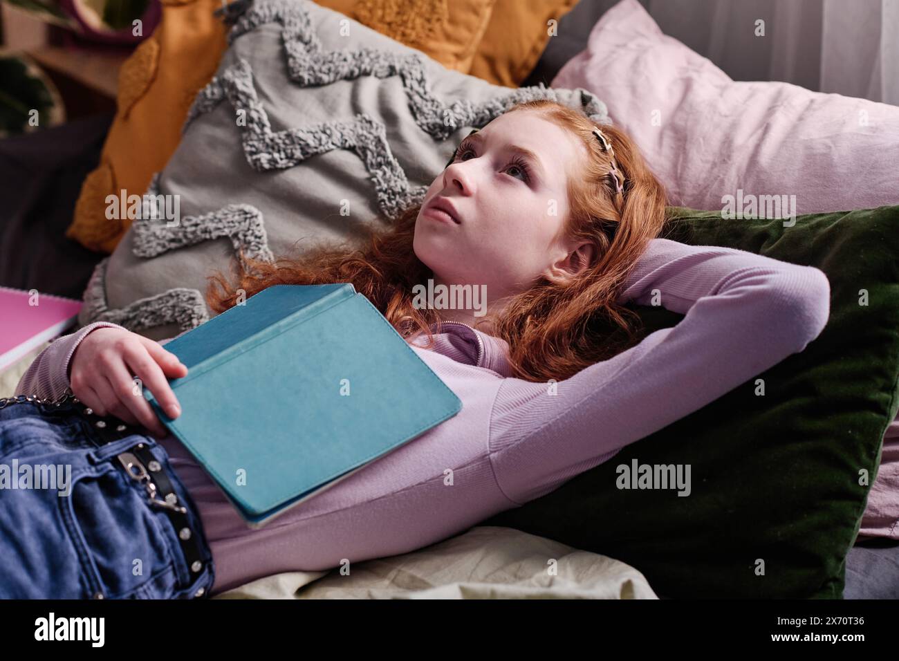 Teen girl holding book relaxing on bed in her bedroom thinking about ...