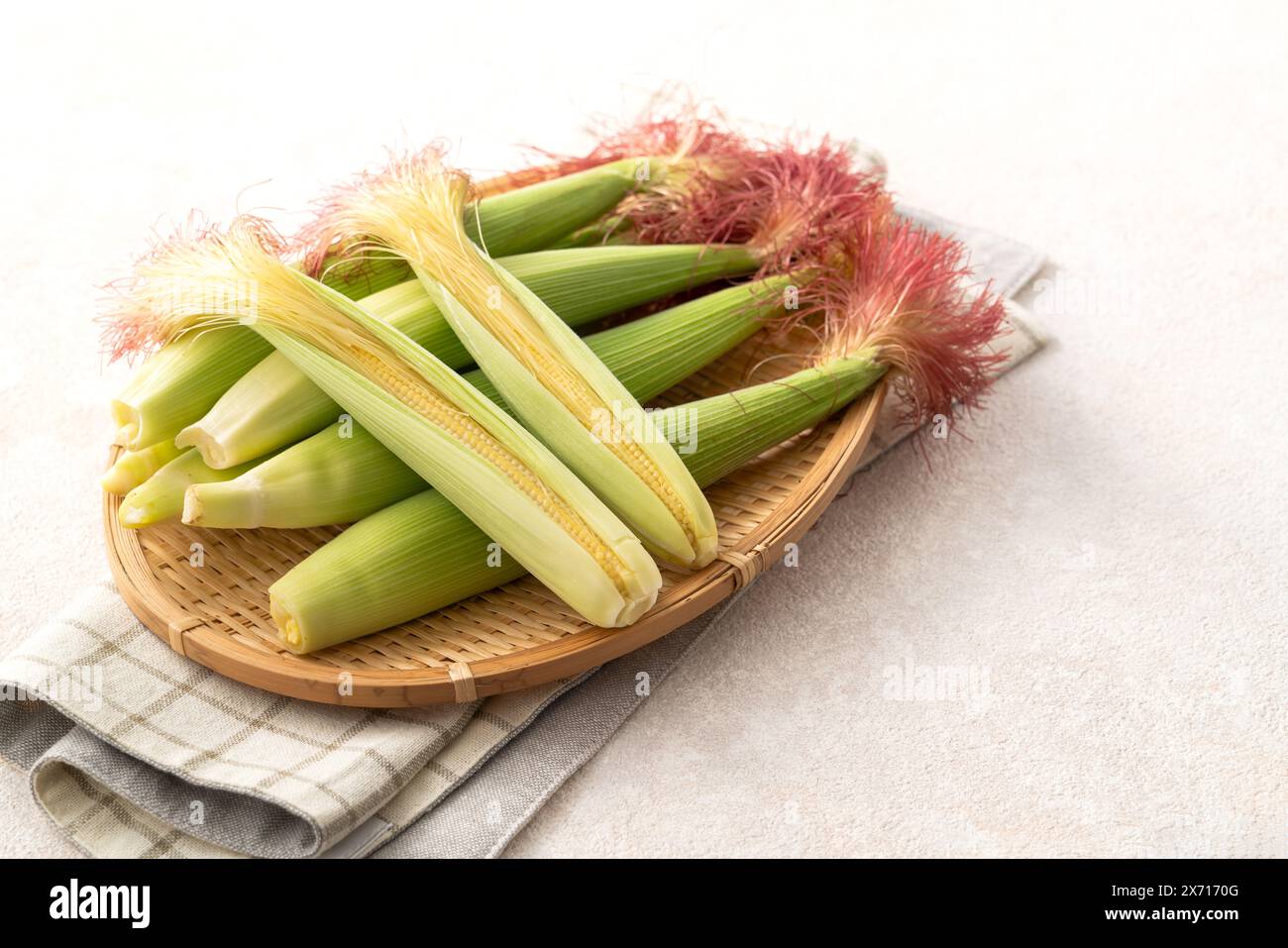 Raw, fresh baby corn in the husk with red silk in basket sieve plate on ...