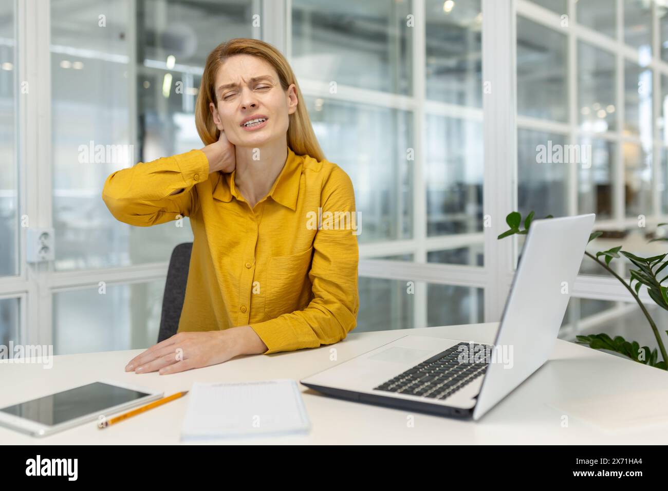 Woman in a yellow shirt feeling neck pain while working on a laptop in ...