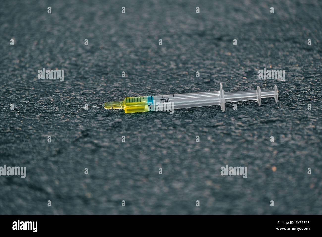 Close-up of a syringe lying on an asphalt road. The syringe contains a ...
