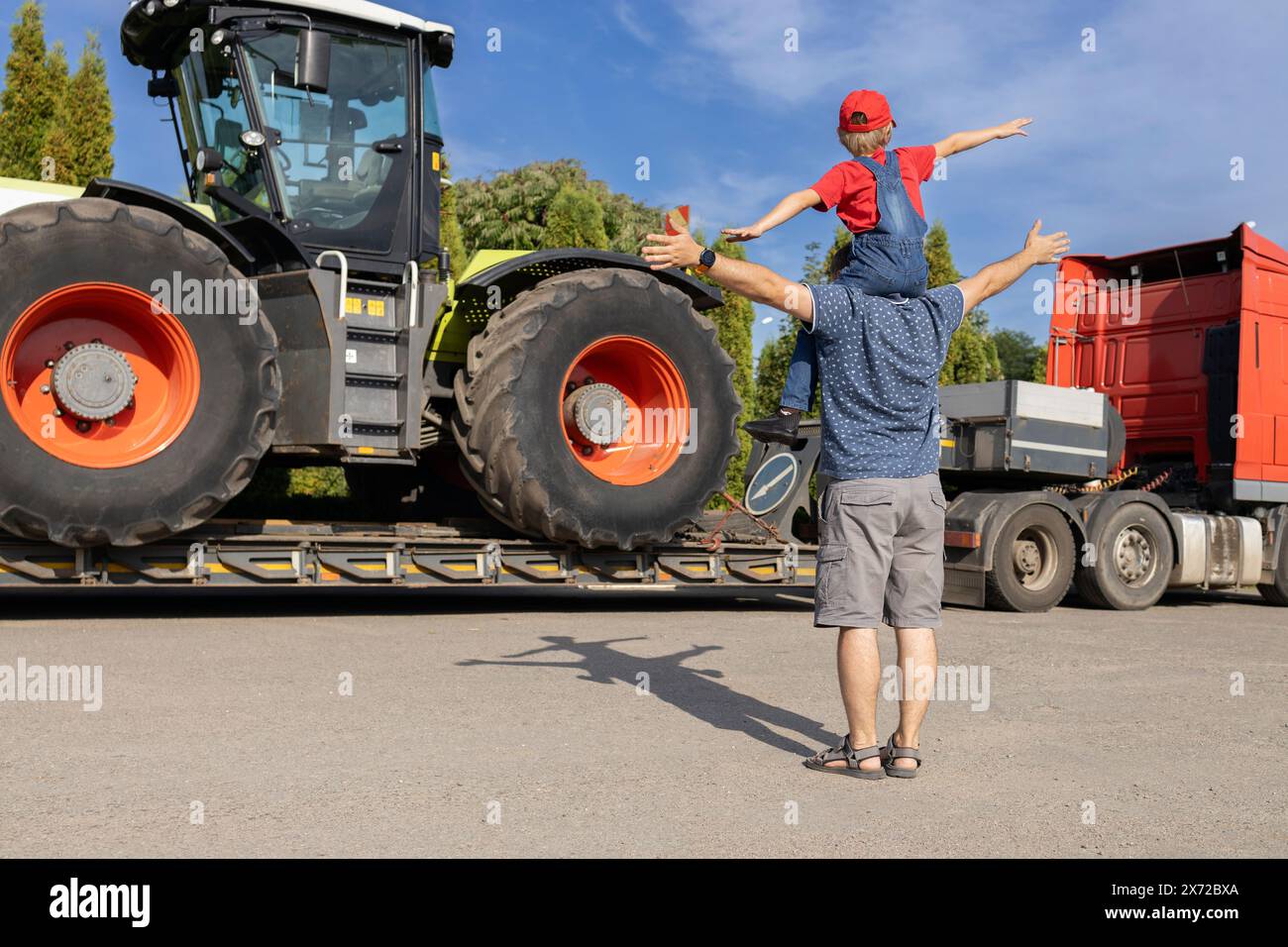 man and boy sitting on his shoulders stand in front of large tractor ...