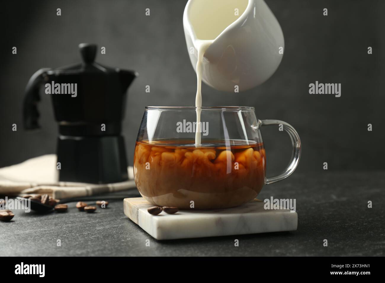 Pouring milk from pitcher into glass cup with coffee at dark textured ...
