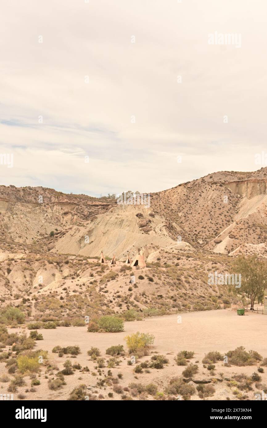 Desert landscape with small teepee structures among rugged hills and sparse vegetation under a ...
