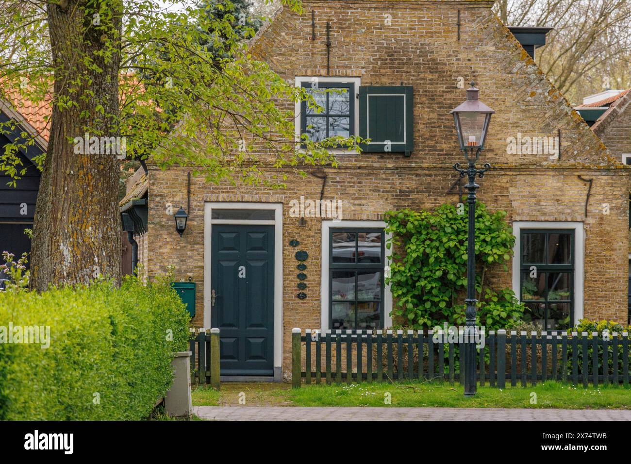 Narrow brick house with garden fence and shutters, flanked by trees ...