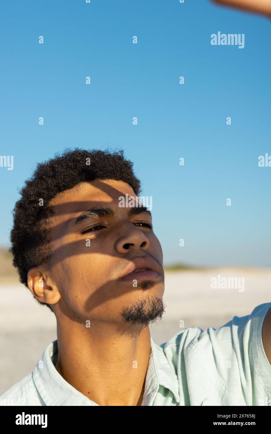 At beach, biracial young man looking at sun, shadows playing on face ...