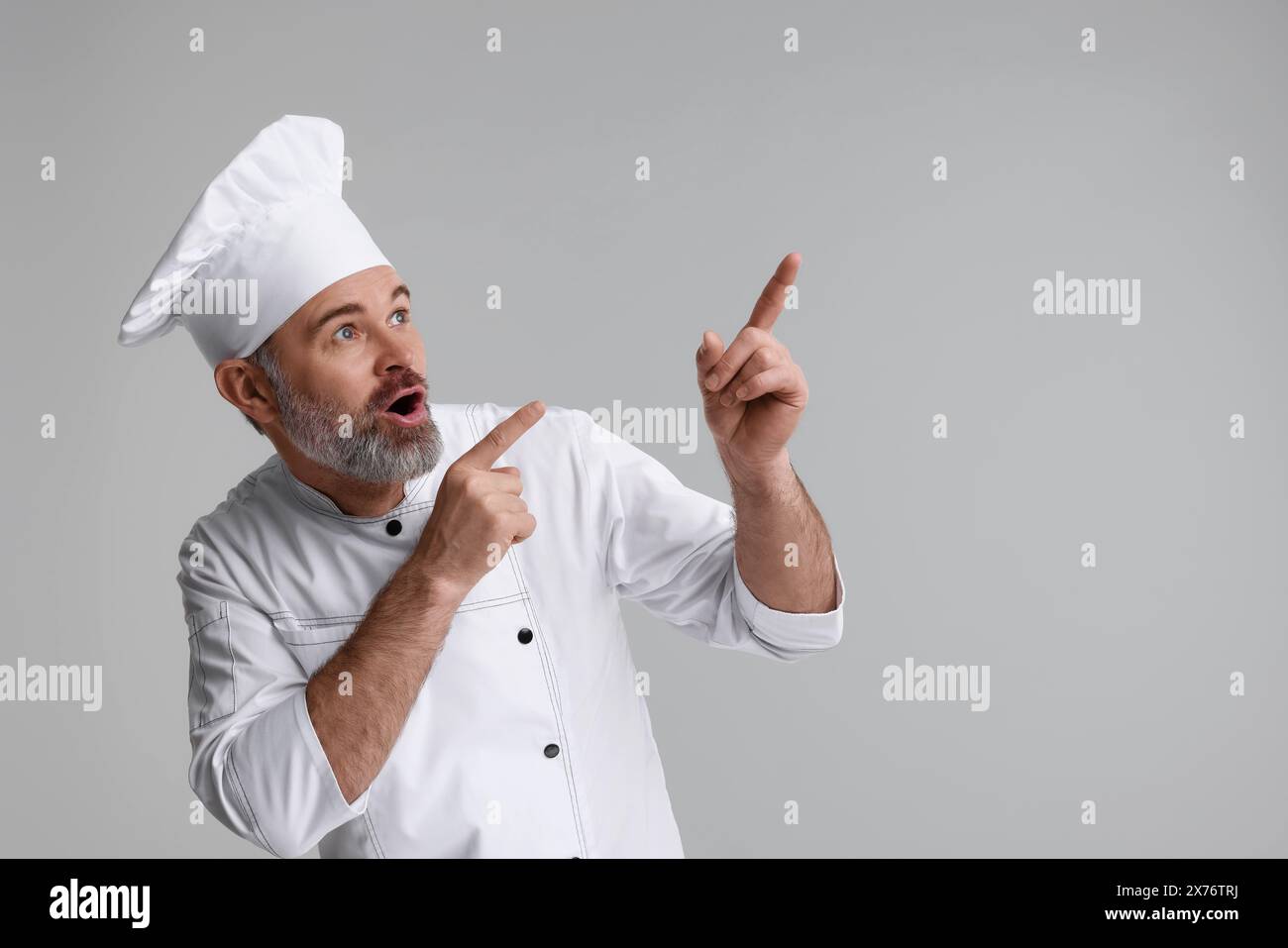 Surprised chef in uniform pointing at something on grey background ...