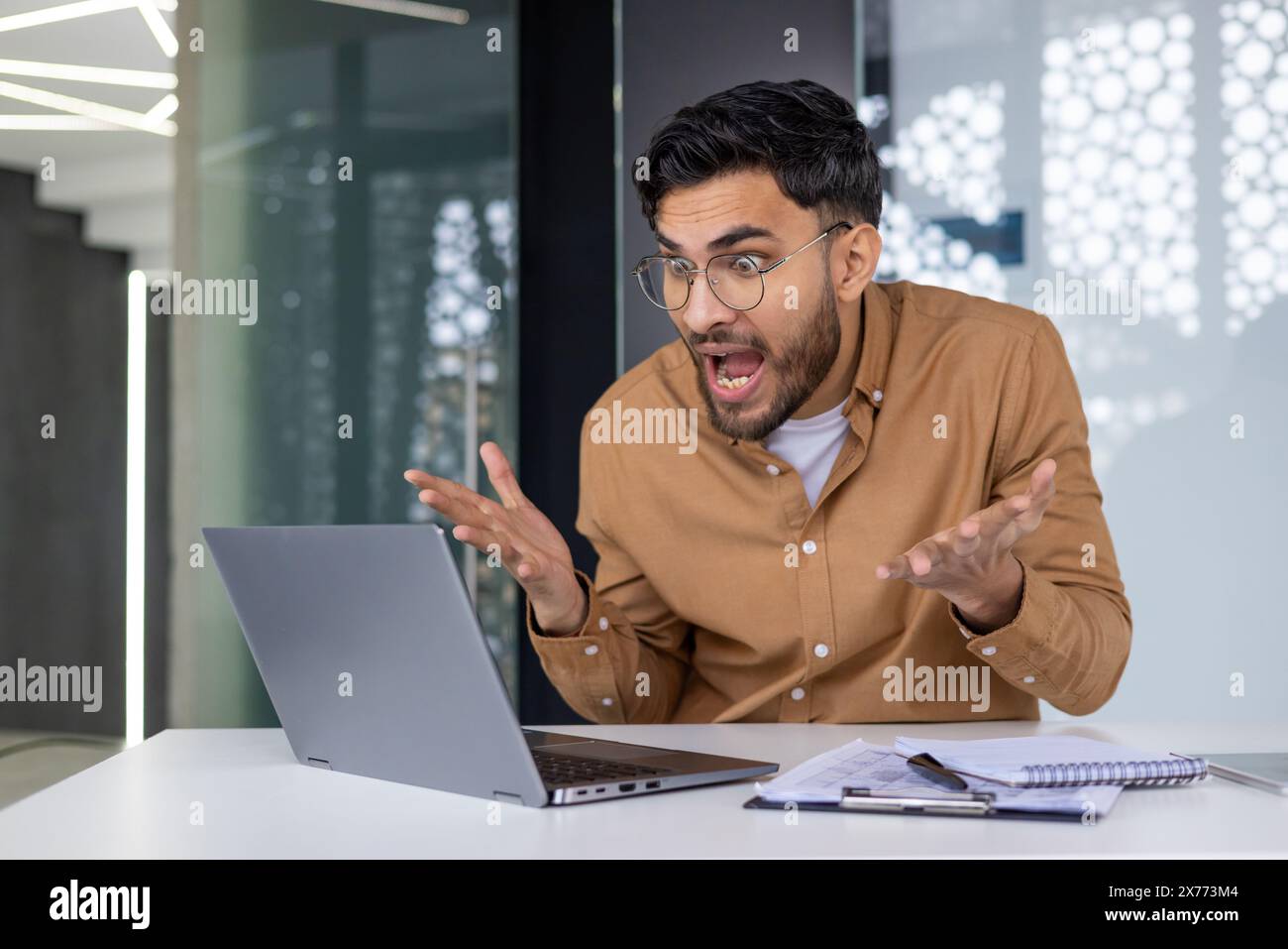 Shocked young man with glasses and beard, gesturing, looking at laptop ...