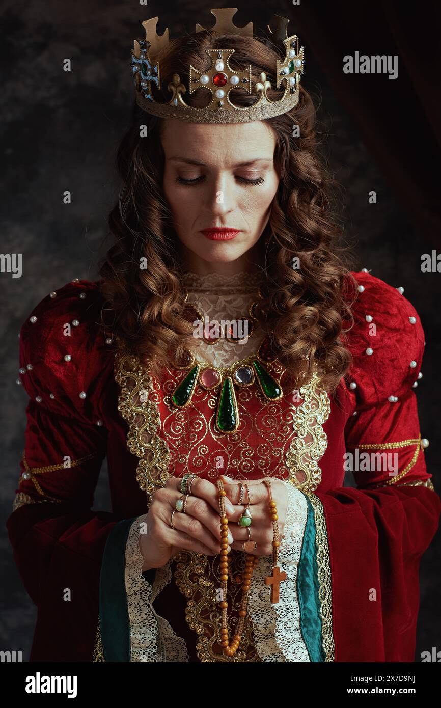 medieval queen in red dress with rosary and crown praying on dark gray ...