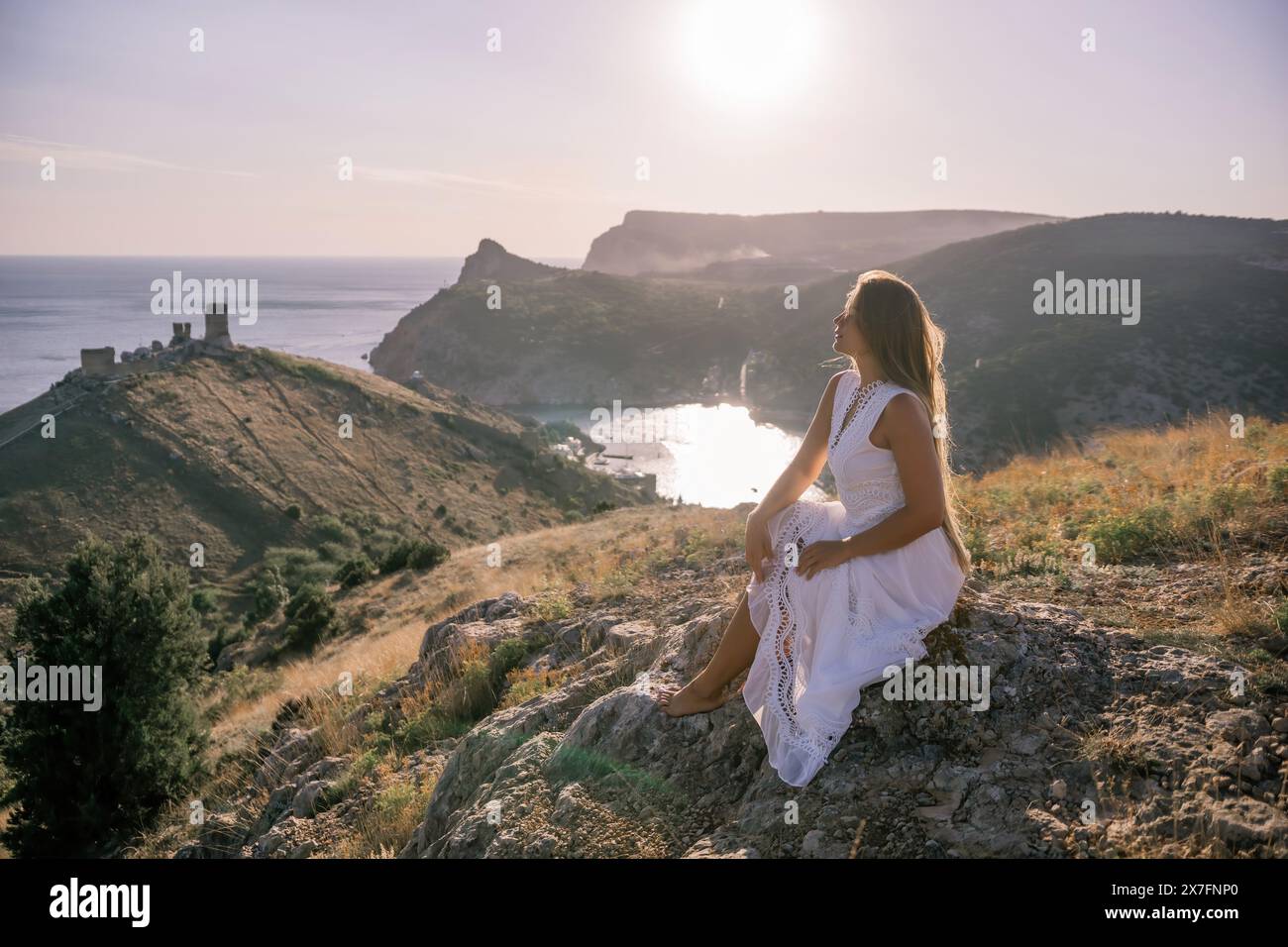 A woman in a white dress sits on a rock overlooking a body of water ...