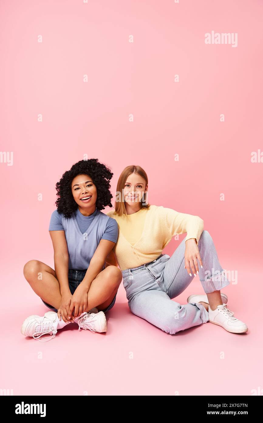 Two diverse women in casual attire sit on the ground smiling for a ...