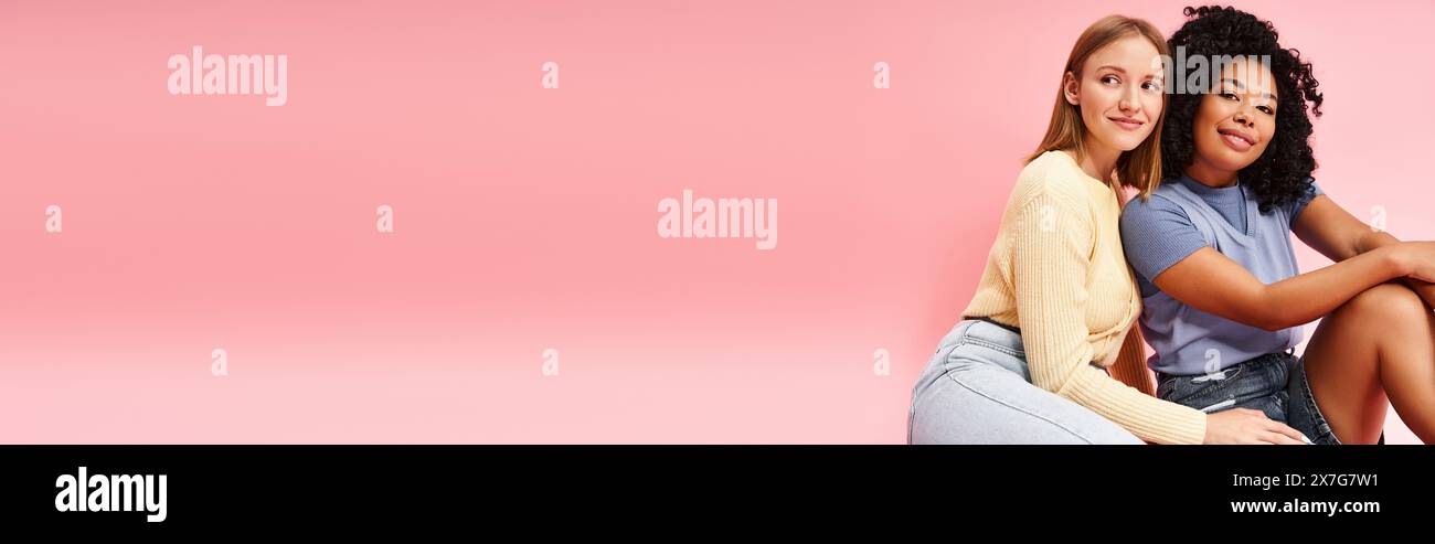 Two attractive diverse women in cozy casual attire sitting on pink ...