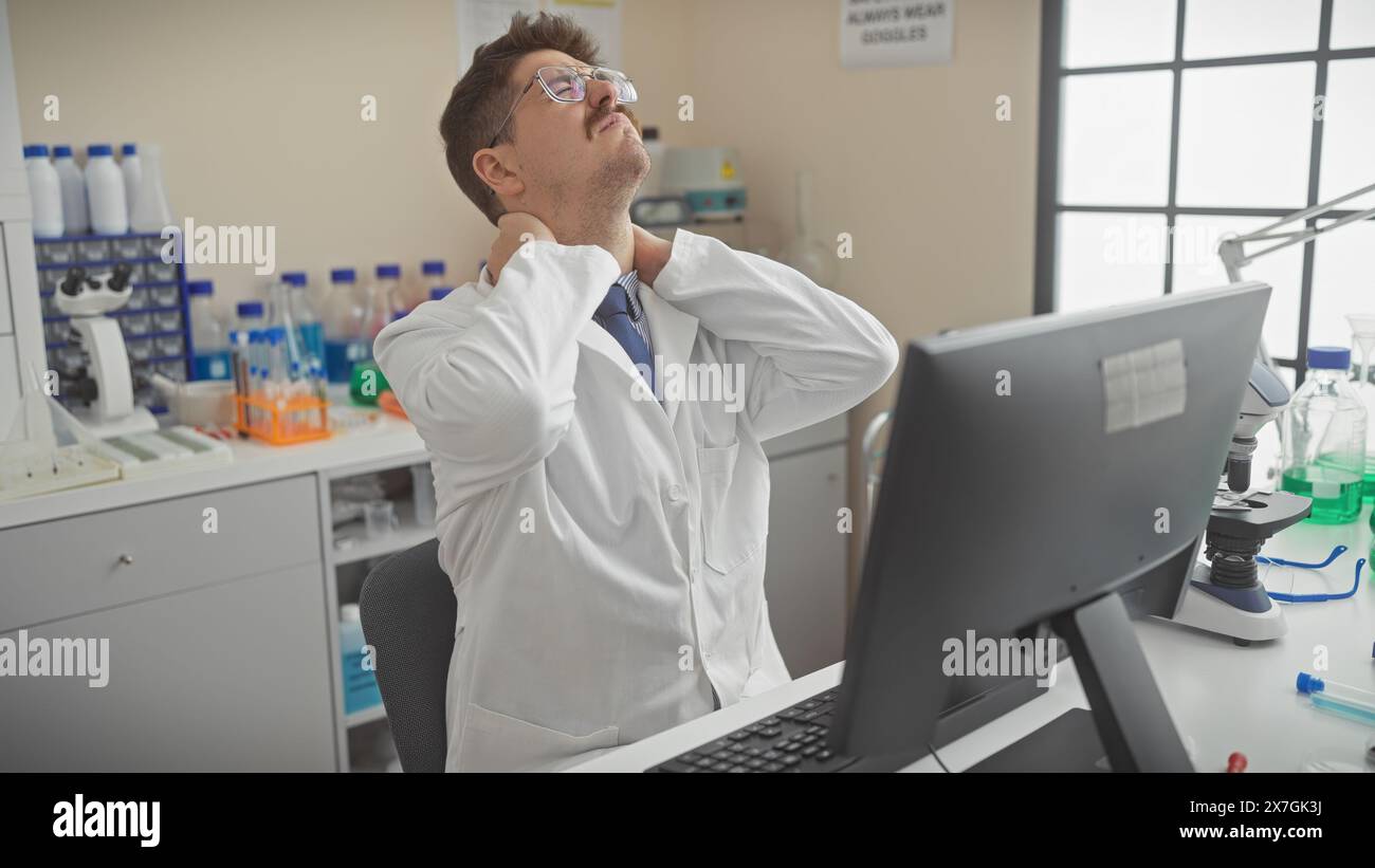 Young man in a lab coat rubbing neck pain at a workstation with ...