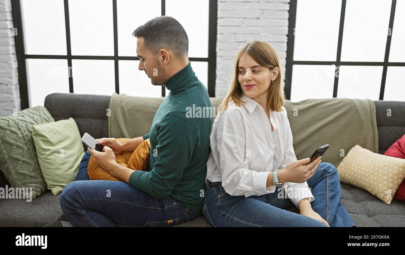 A woman sneakily glances at a man's smartphone as they sit back-to-back on a couch indoors Stock ...