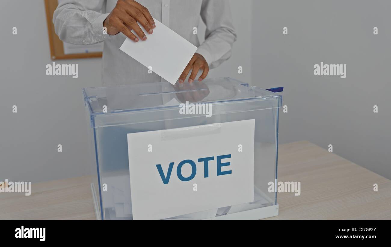 A young adult man is placing a ballot in a transparent box at an indoor ...