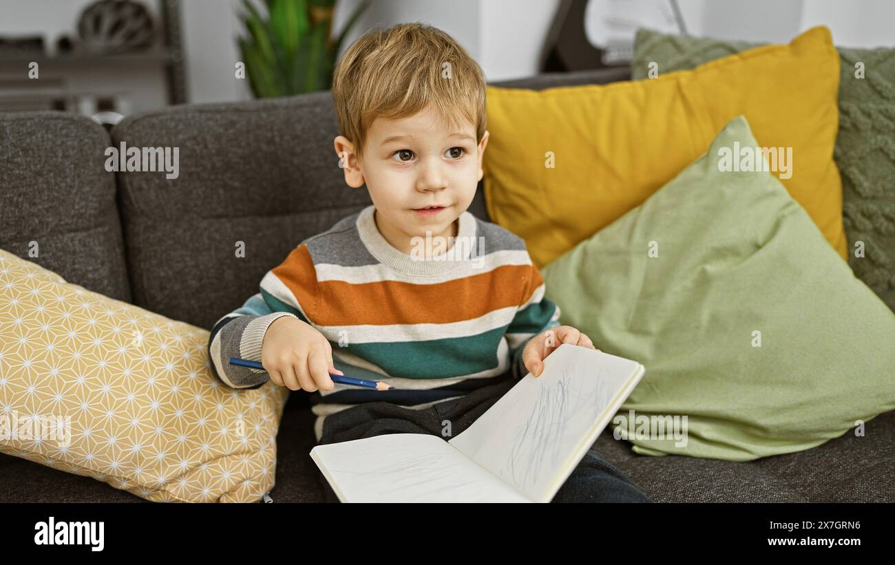 Calm toddler boy with blond hair drawing in a notebook on a gray couch ...
