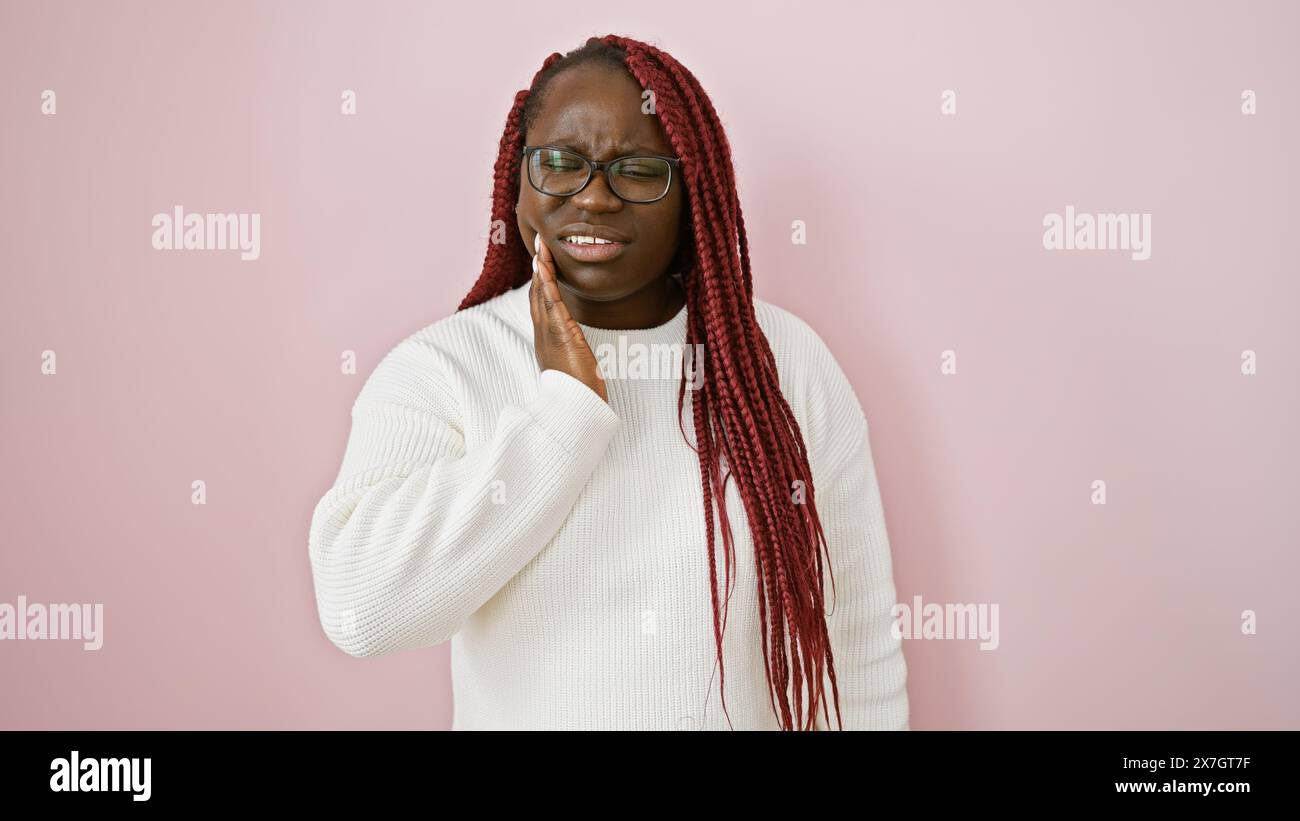 A pained african woman with braids touches her cheek against a pink ...