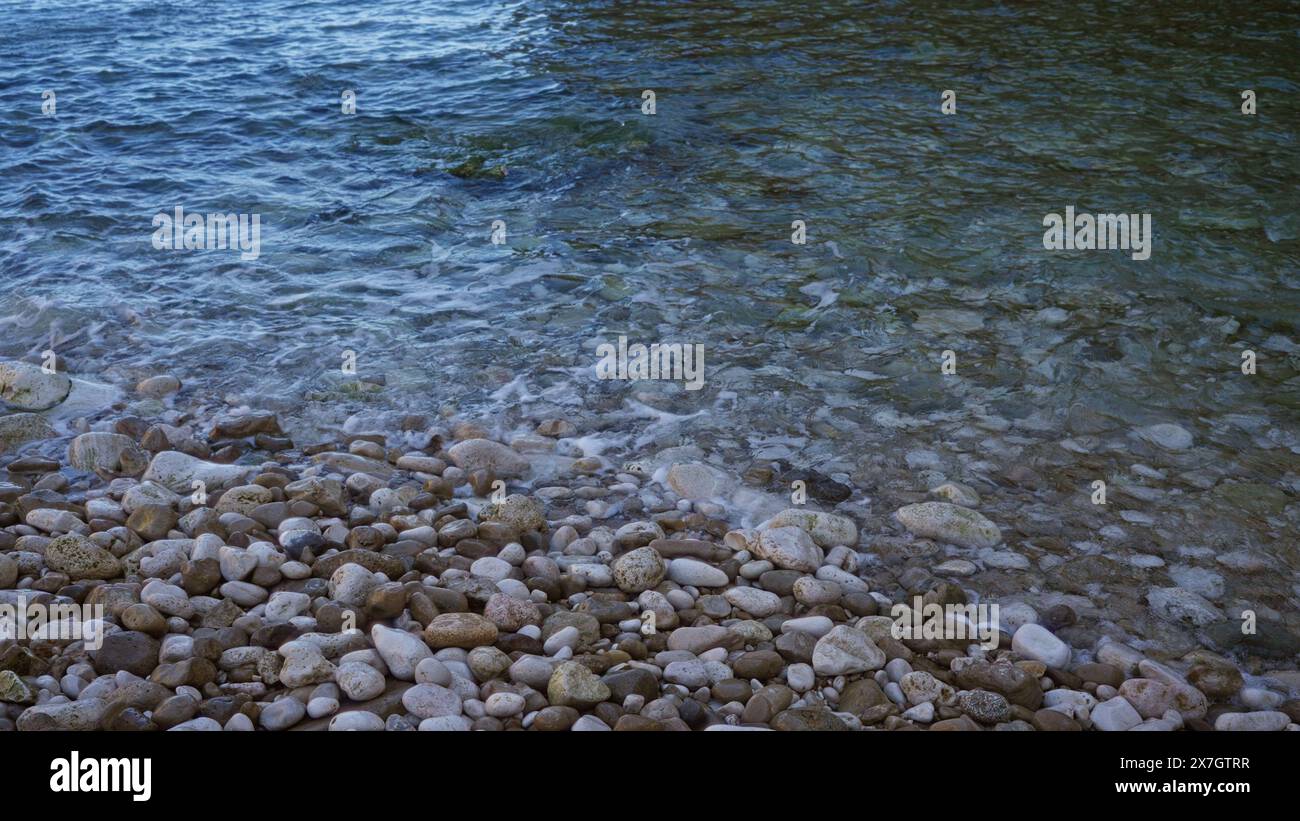 A serene pebbled beach shoreline with clear blue water and gentle waves ...