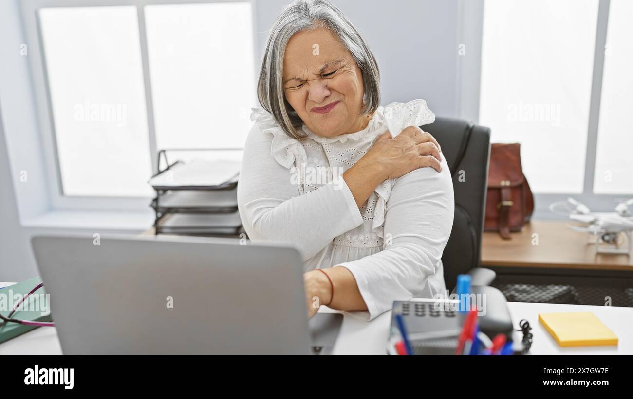 Mature grey-haired woman grimacing in pain at office due to shoulder ache Stock Photo - Alamy