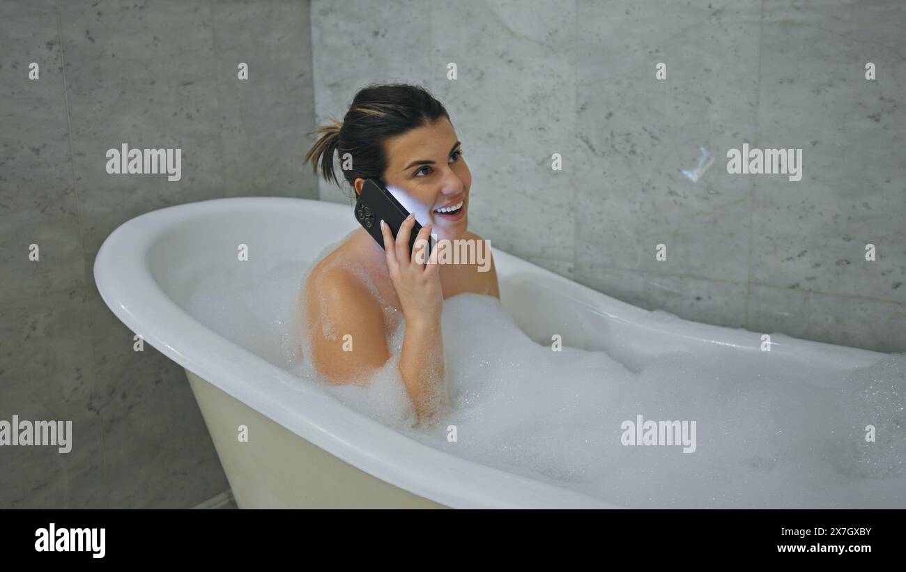 A joyful young woman chats on a smartphone while relaxing in a foamy ...