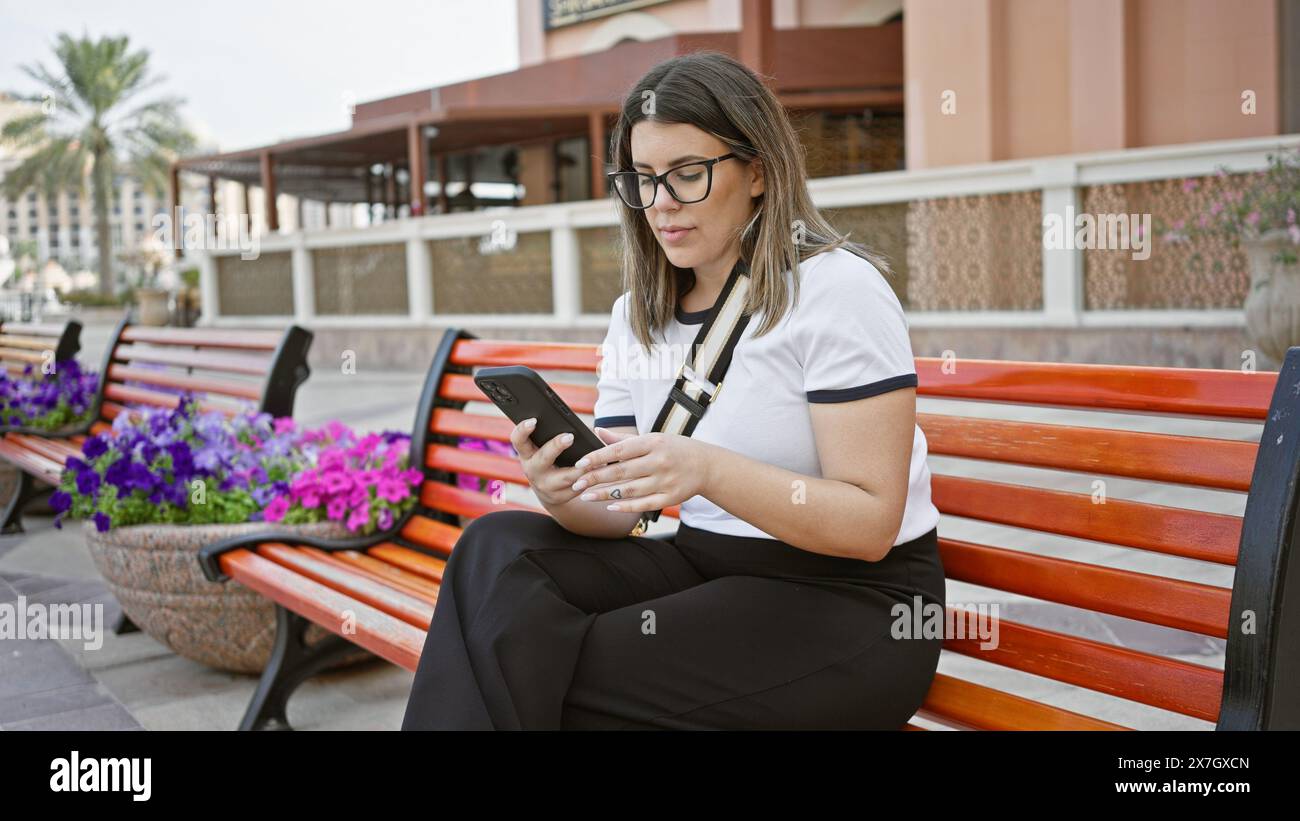 A young adult woman checks her phone while seated on a vibrant street ...
