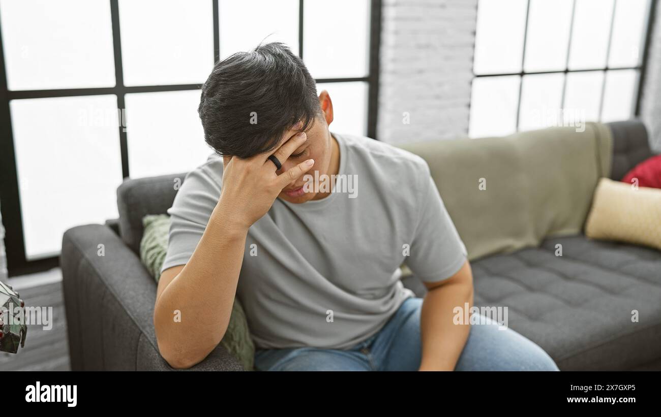 A stressed asian man sitting on a sofa indoors, covering face with hand ...