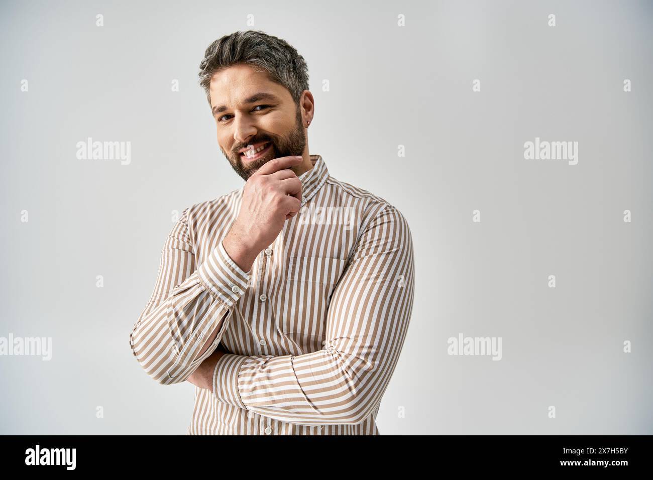 A stylish man with a beard poses in elegant attire against a grey ...