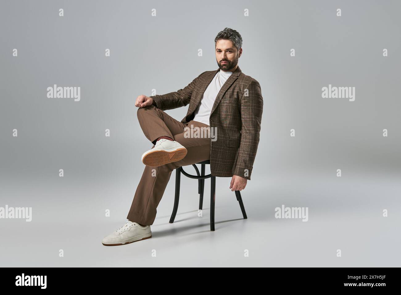 A stylish man with a beard sits on a chair, crossing his legs elegantly ...