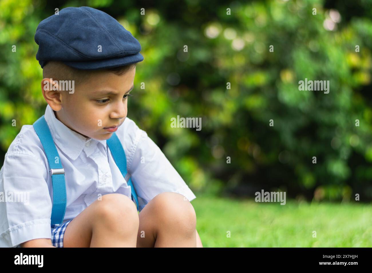very sad little latino boy sitting on the ground in a public park ...