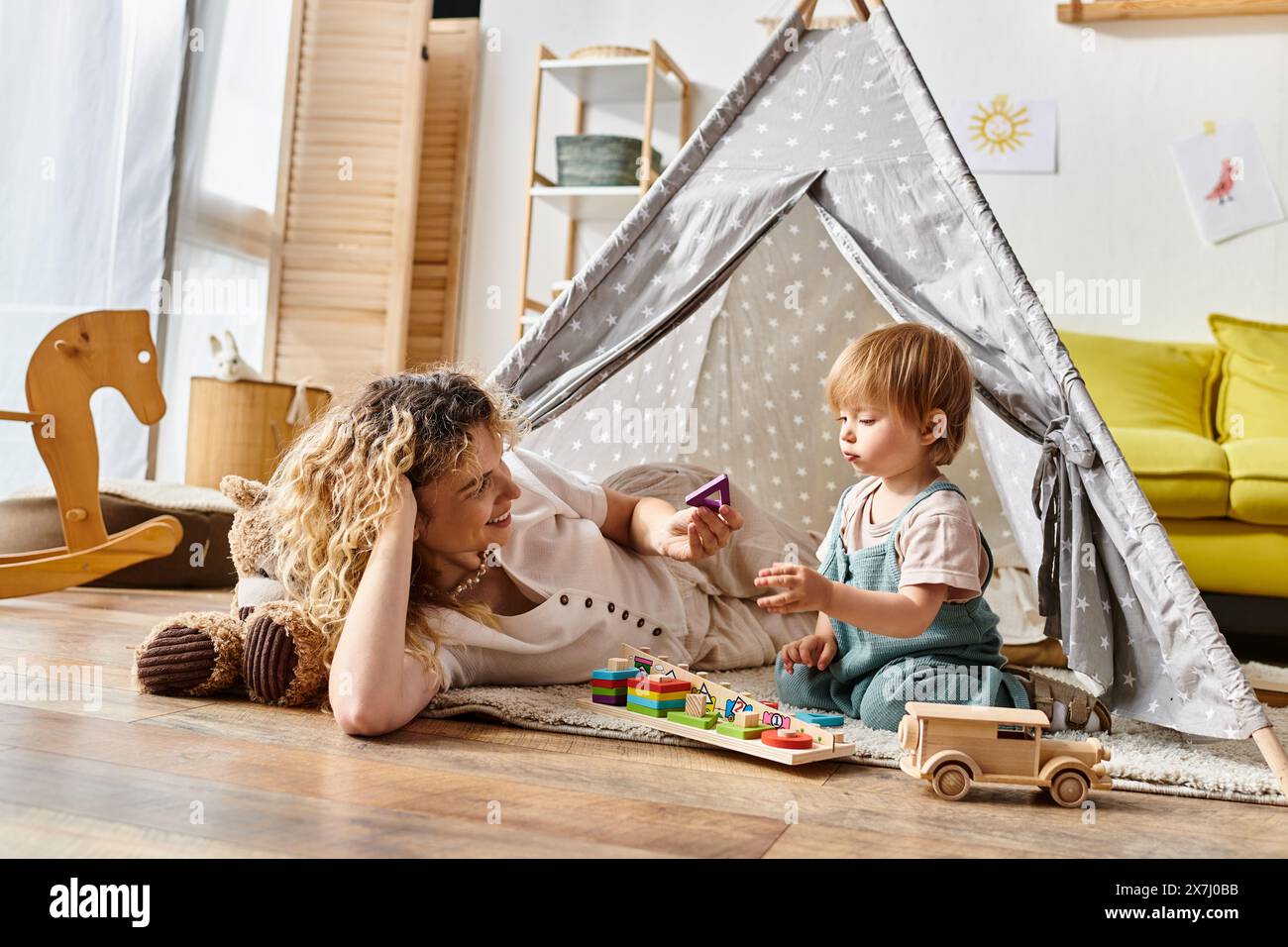 Curly mother and toddler daughter happily playing together inside a colorful play tent ...