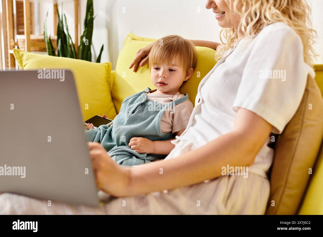 A curly-haired mother sitting on a couch, tenderly holding her toddler daughter on her lap in a ...