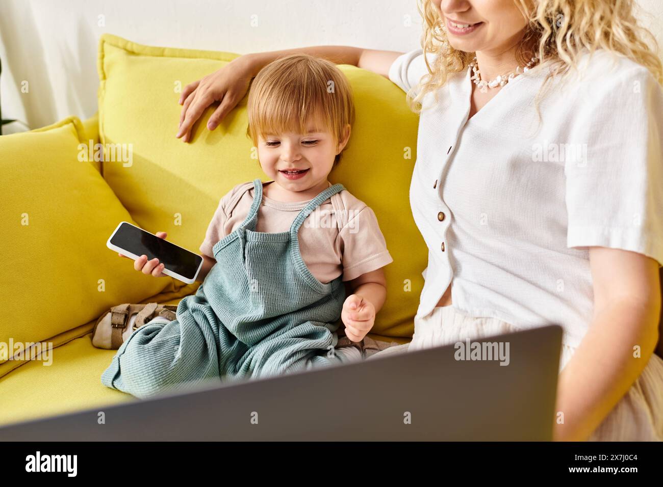 Curly mother and her toddler daughter sitting on a couch, engrossed in ...