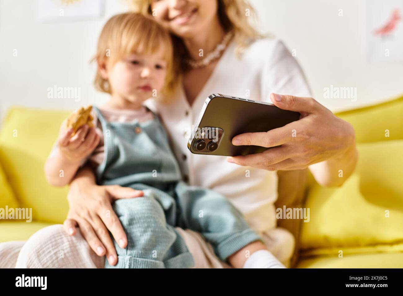 A curly mother holds a cell phone next to her toddler daughter at home ...