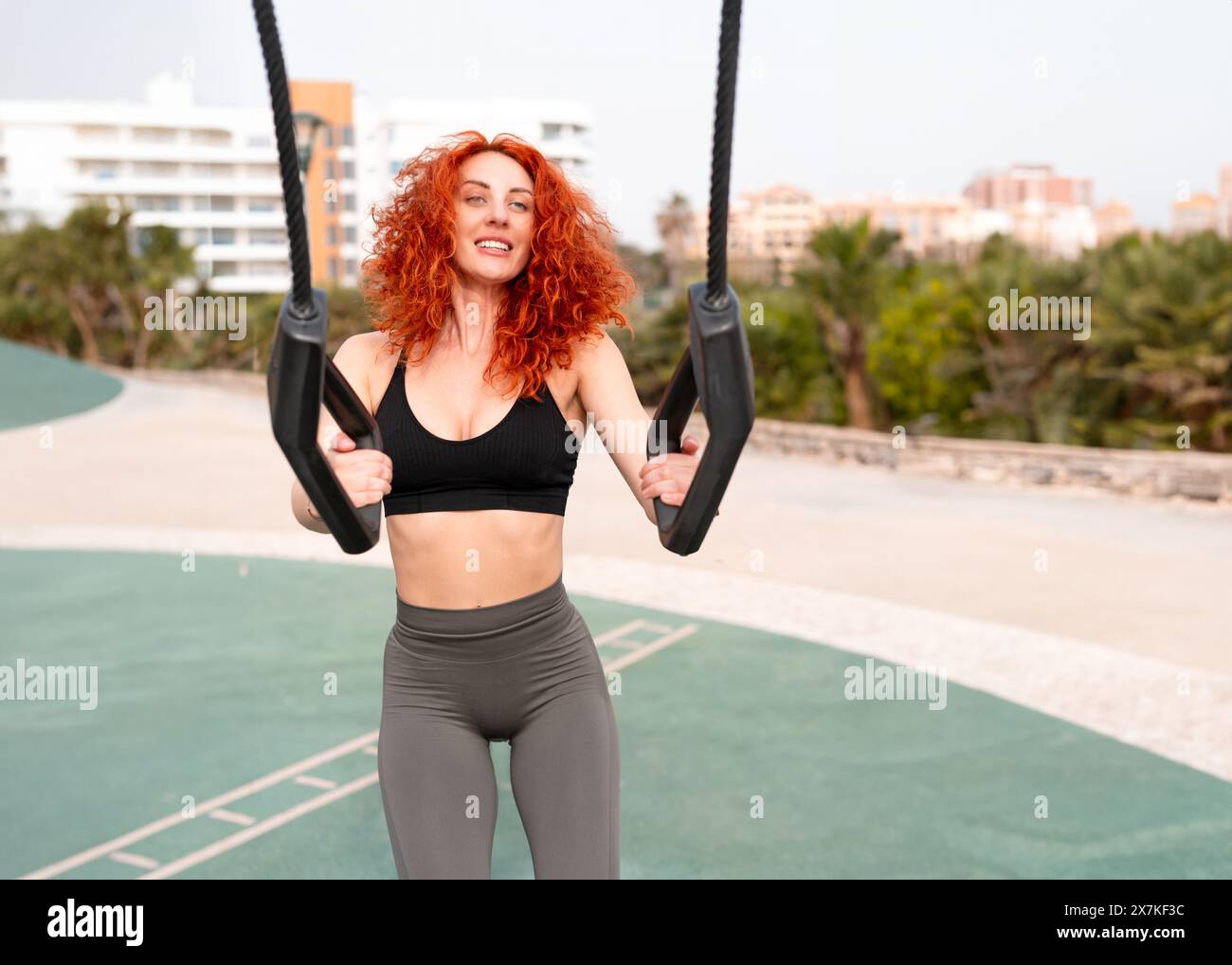 Happy slim woman doing suspension training with fitness straps outdoors. Fit strong redhead ...