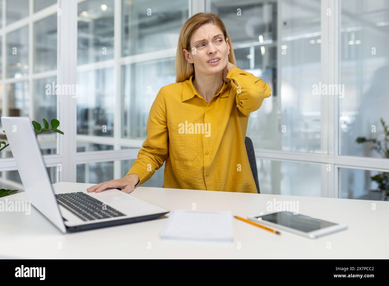 Woman in yellow shirt experiencing neck pain while working at her desk ...