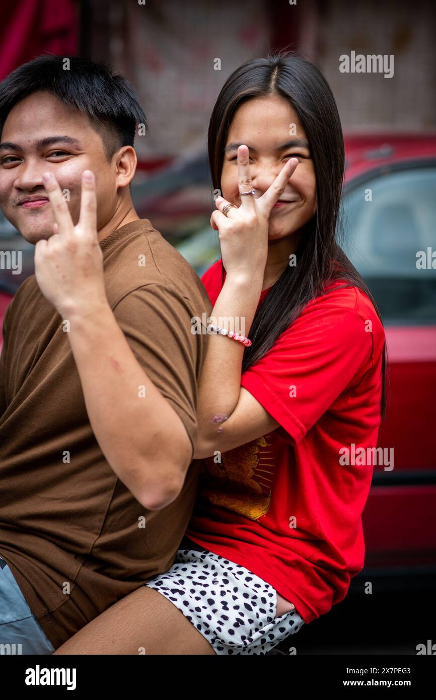 A young Filipino couple speed by on a scooter and make fun hand signals ...