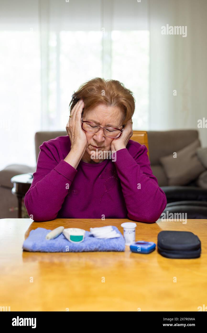 A senior woman sits at a table feeling distressed and holding her head ...