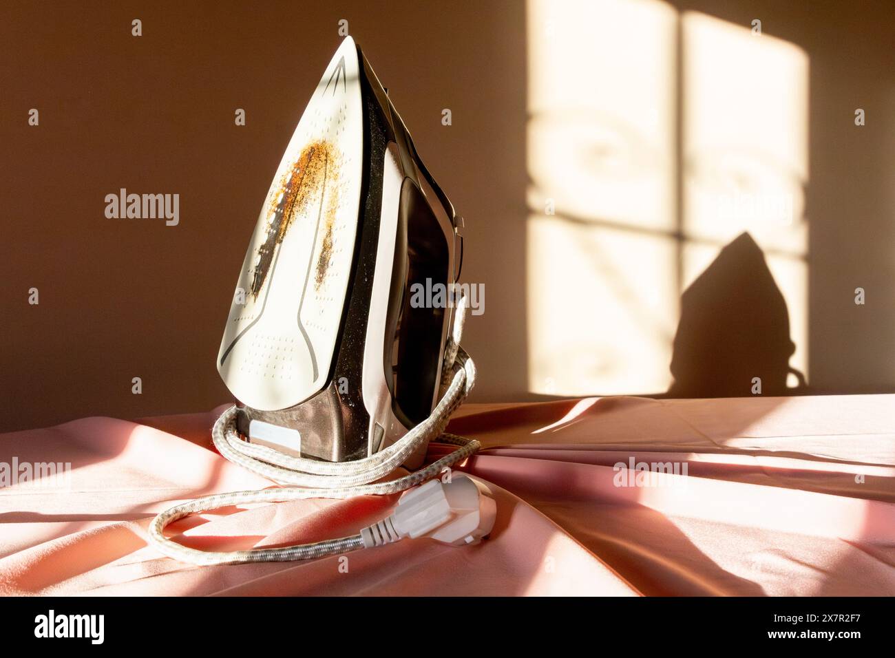 A steam iron with a scorched soleplate rests on pink fabric, bathed in ...