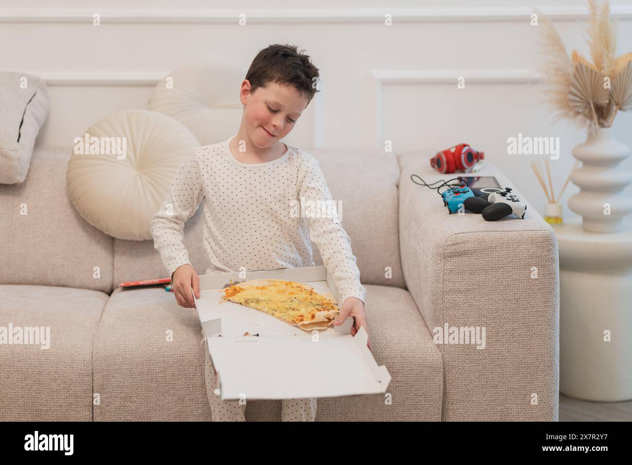 A cheerful boy sits on a couch holding a pizza box, smiling down at a ...