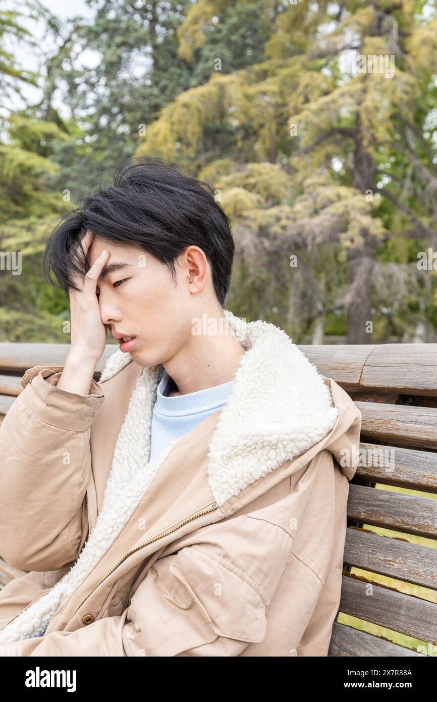A young Asian man sits on a park bench, pressing his hand to his ...