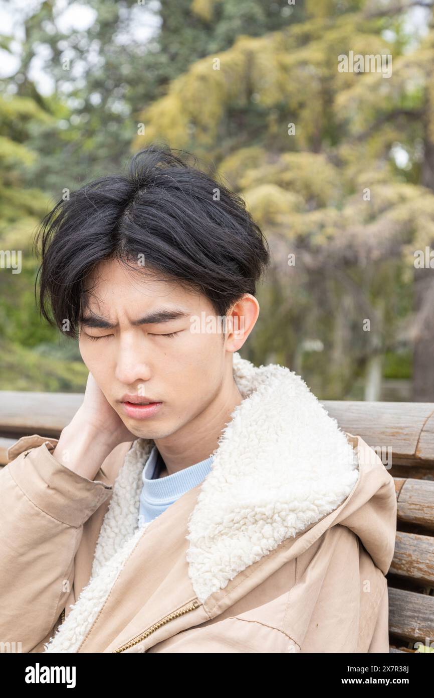 A young Asian male sitting outdoors with a pained expression, clutching ...