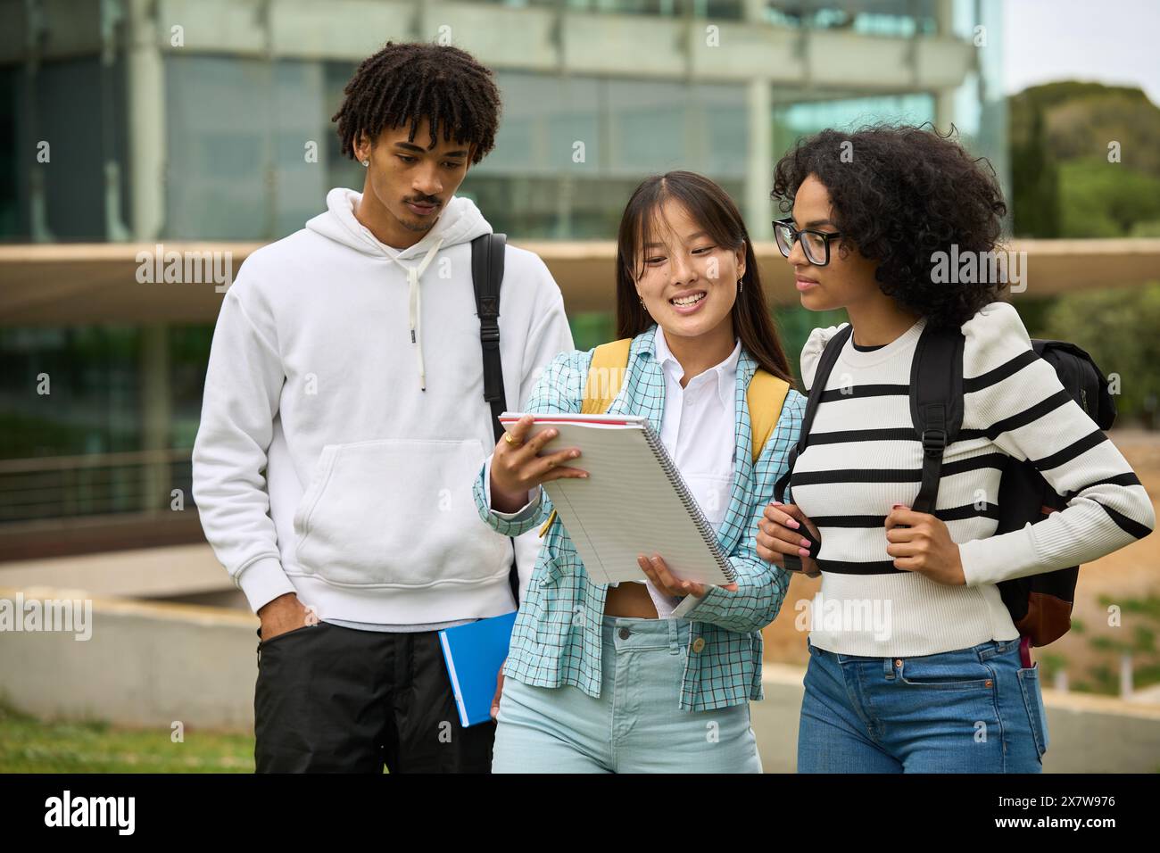 Three young students are standing outside a building sharing notes ...