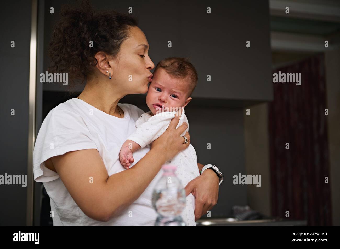 Affectionate mother kissing the temple of her child's head, measuring the temperature of her ...