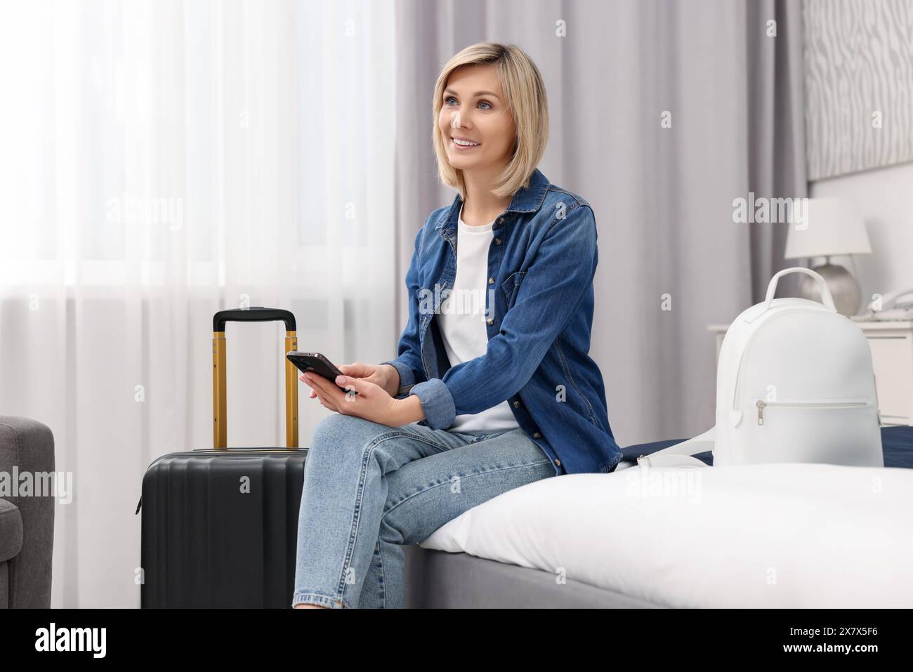Smiling guest with smartphone relaxing on bed in stylish hotel room ...