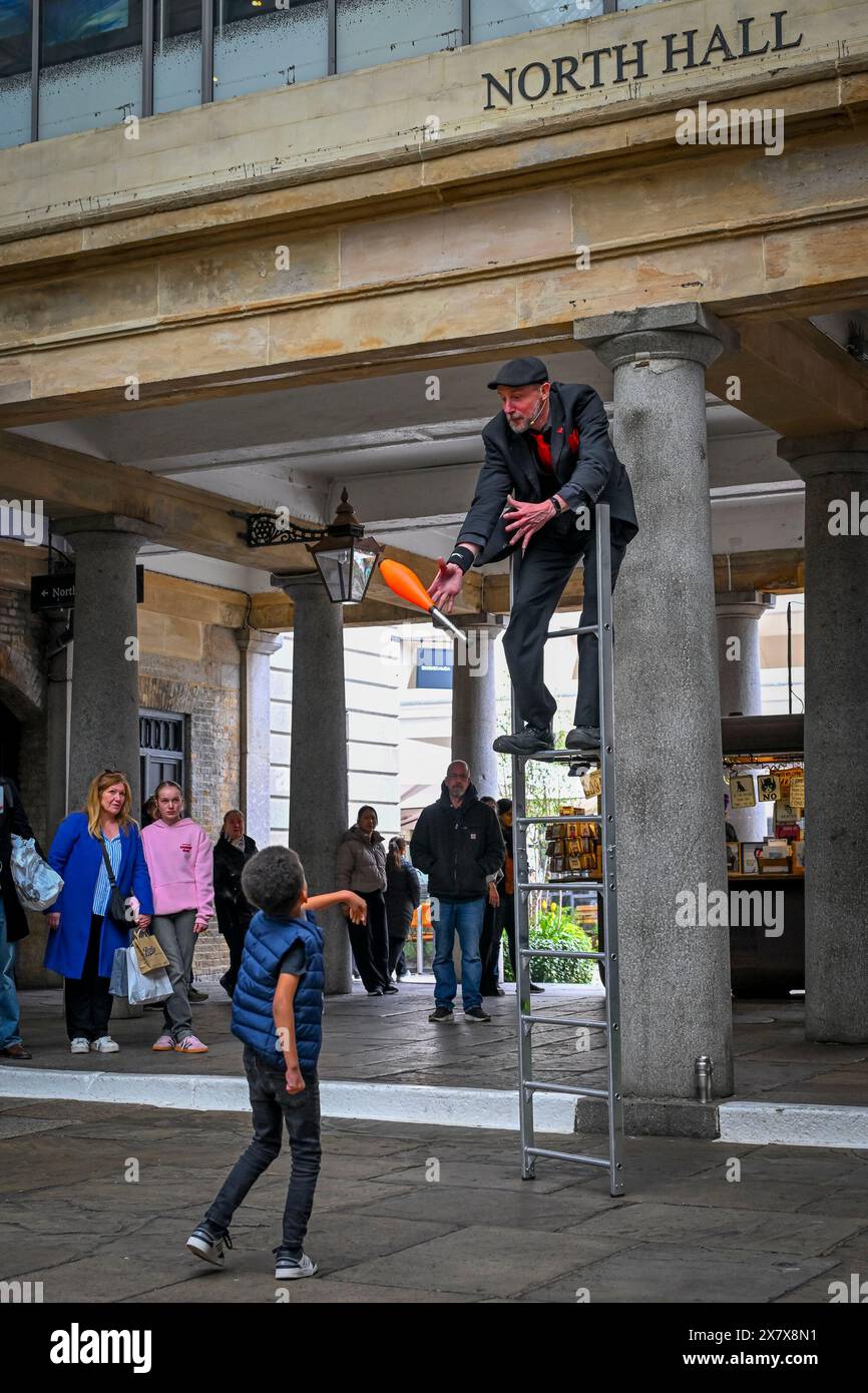 Busker balancing on ladder, while young boy throws him a juggling pin ...